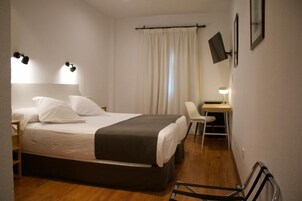 Quarto com cama, mesa de cabeceira, cadeira, escrivaninha e televisão na parede.