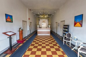 Um saguão de hotel com balcão de recepção, mesa de atendimento, área de estar com uma cadeira, uma pequena mesa e uma estante de livros.