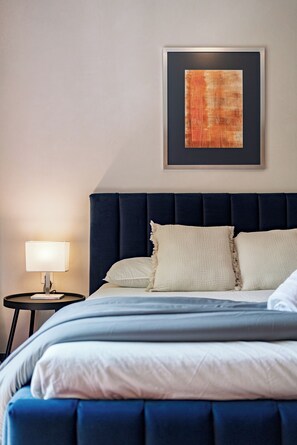 Uma cama bem arrumada com cabeceira escura, abajur de cabeceira e uma pintura abstrata emoldurada na parede.