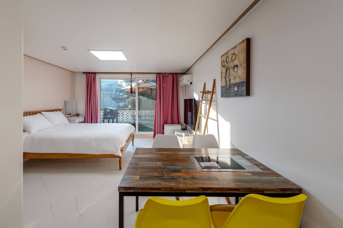 Quarto com cama, escrivaninha, cadeiras, um quadro e varanda com cortinas.