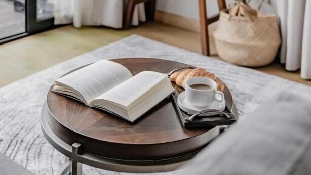 Um cantinho aconchegante para leitura, com uma mesa redonda de madeira, um livro, uma xícara de café e um croissant.