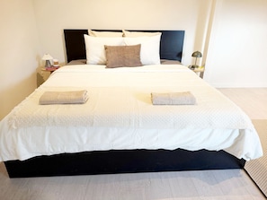 Cama bem arrumada com cabeceira preta, roupa de cama branca e duas toalhas dobradas.