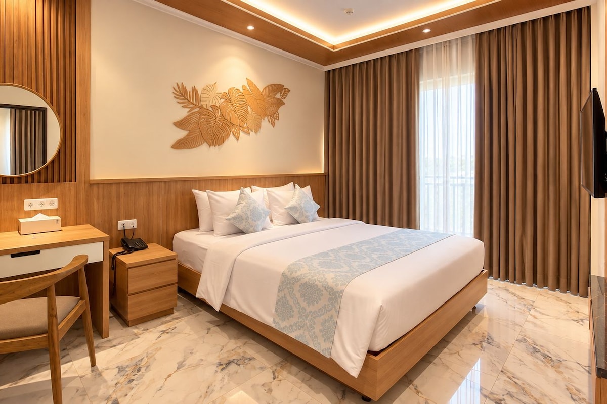 Quarto de hotel com cama, escrivaninha, cadeira, mesa de cabeceira, espelho e televisão fixada na parede.
