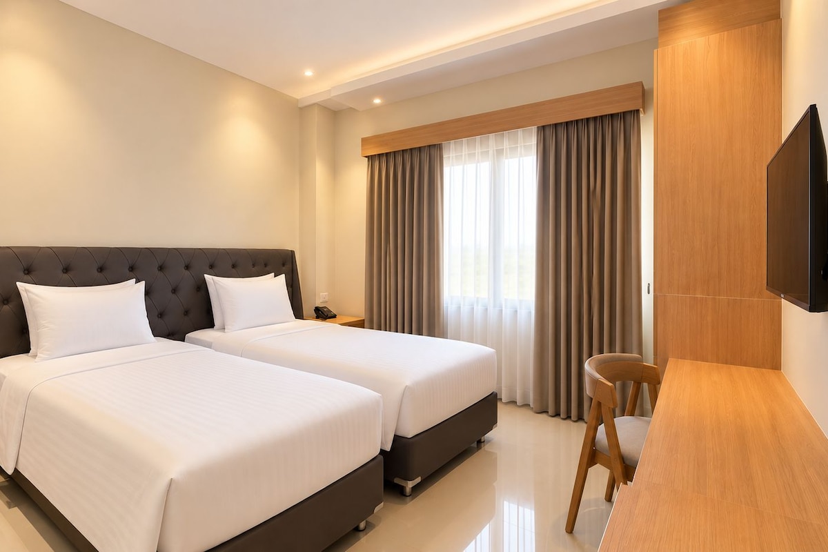 Quarto de hotel com duas camas, uma escrivaninha, uma cadeira, uma televisão e uma janela com cortinas.