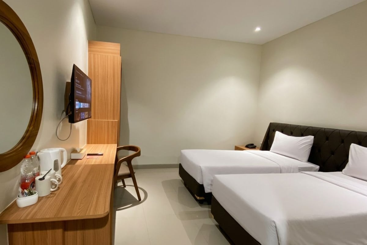 Quarto de hotel com duas camas, uma escrivaninha com cadeira, um espelho grande e uma televisão na parede.