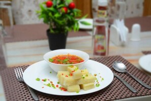 Um prato com comida e uma tigela, um garfo, uma colher e um vaso de planta ao fundo.
