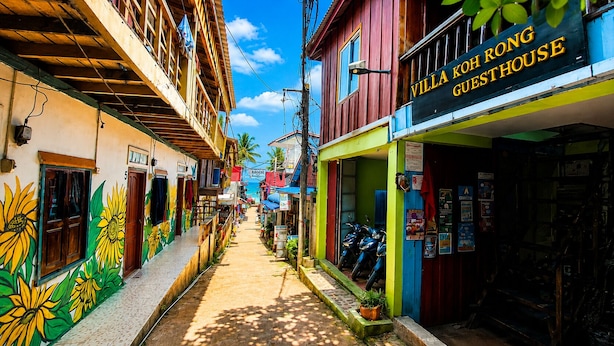Uma rua estreita com edifícios coloridos, uma pensão chamada 'Villa Koh Rong Guesthouse' e uma moto estacionada na frente.