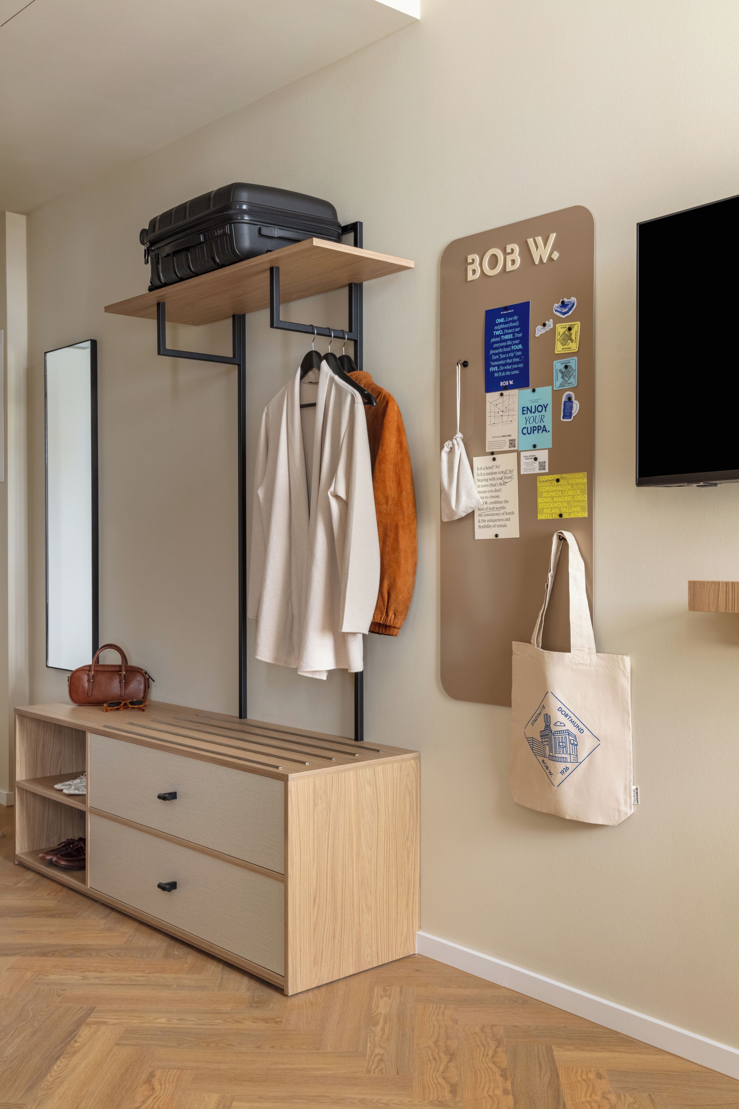 Quarto de hotel com uma mesa de consola de madeira, cabideiro suspenso, TV fixada na parede e um mural com notas sobre viagens.