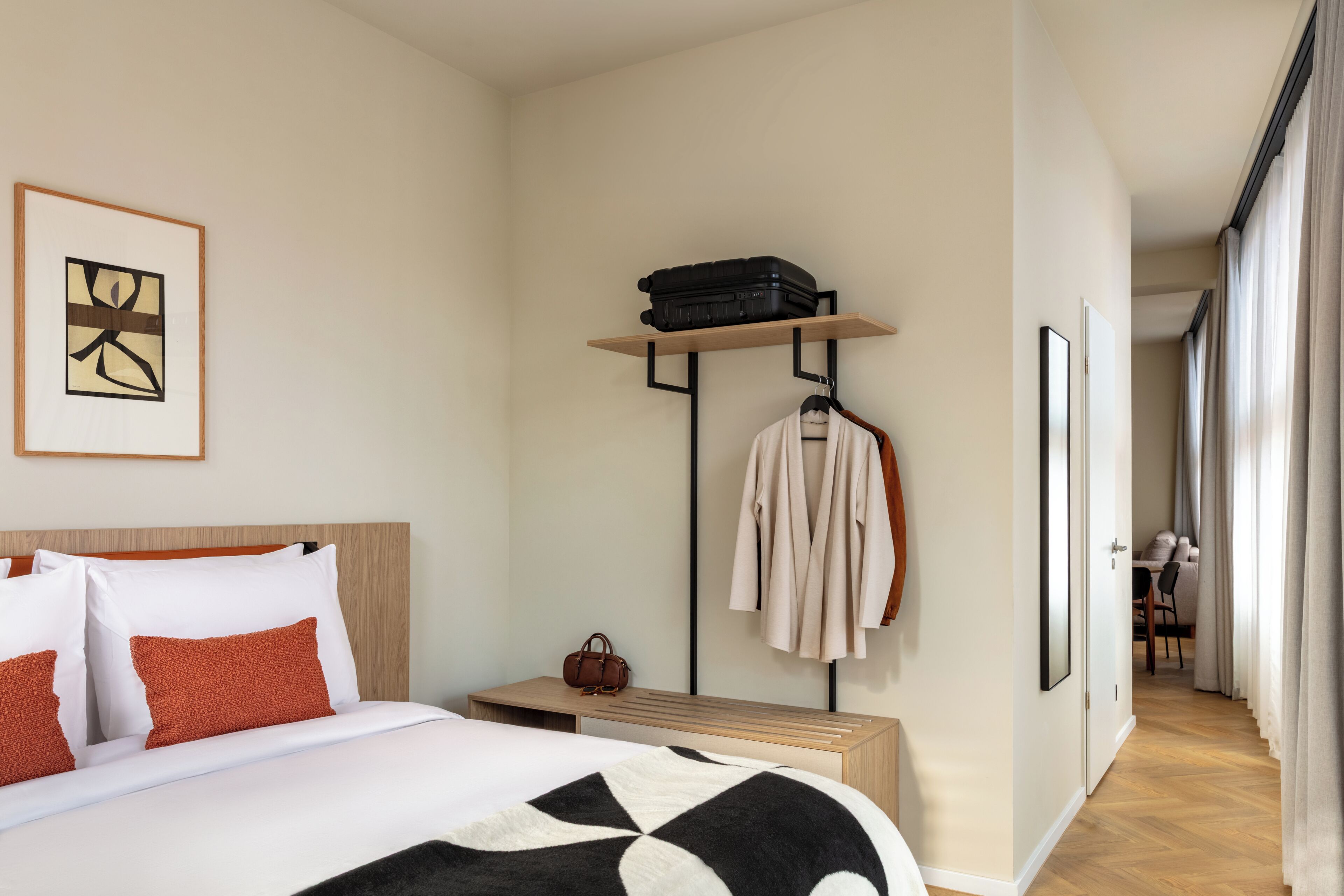 Quarto de hotel com uma cama, mesa de cabeceira, cabideiro e mala sobre uma prateleira.