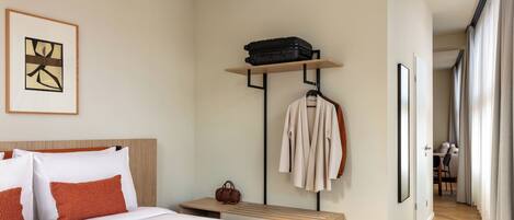 Quarto de hotel com uma cama, mesa de cabeceira, cabideiro e mala sobre uma prateleira.