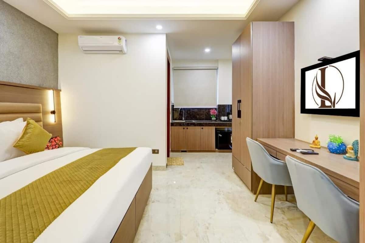 Quarto de hotel com cama, escrivaninha, cadeira, pequeno espaço de cozinha e obra de arte na parede.