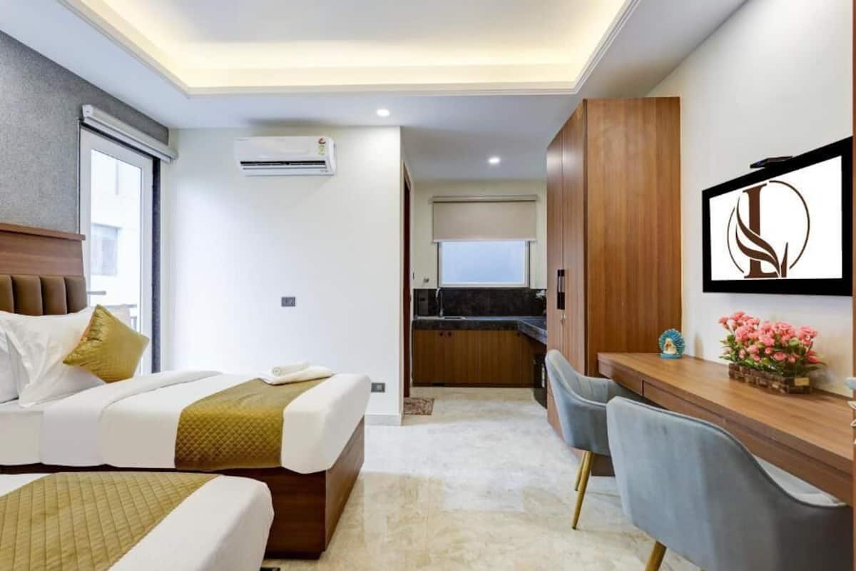 Quarto de hotel com cama, escrivaninha, cadeira, cozinha compacta e televisão.