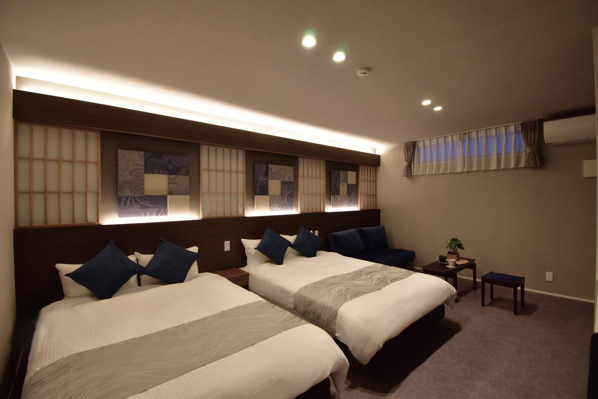 Quarto de hotel com duas camas, uma mesa pequena, uma cadeira e um vaso de planta.