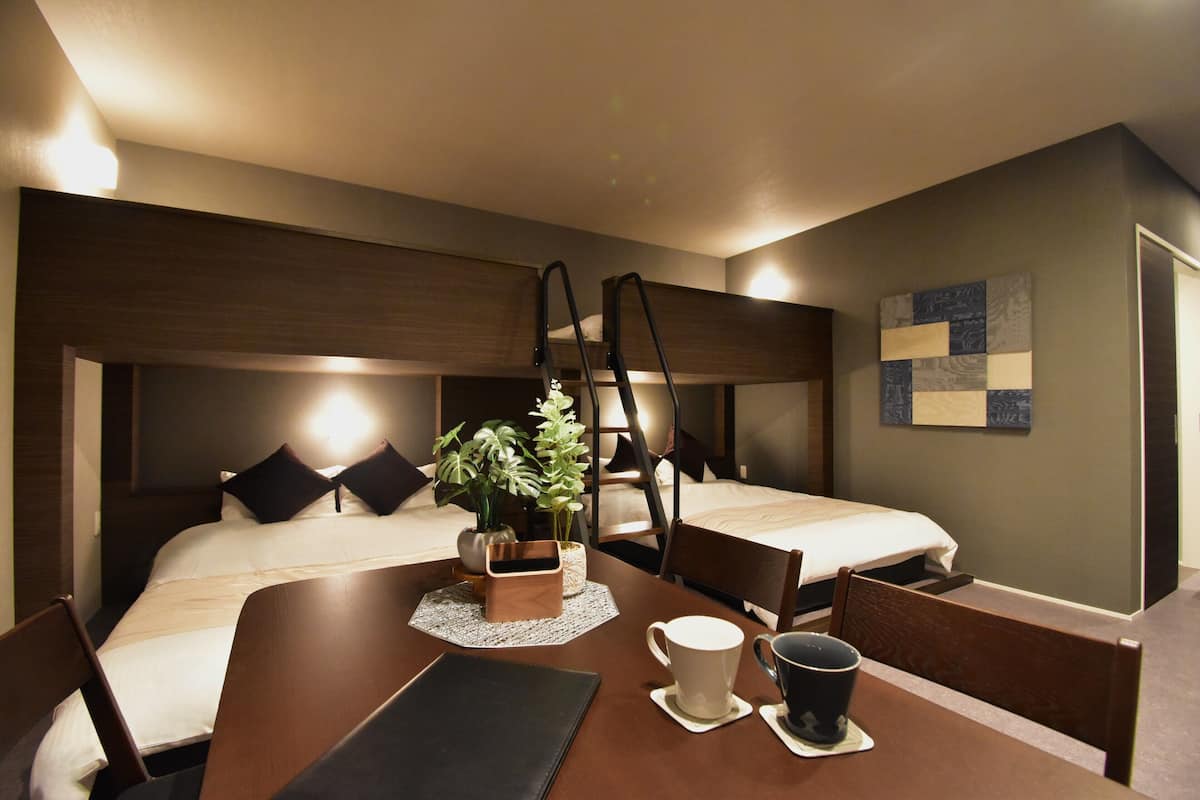 Quarto de hotel com duas camas, uma mesa de jantar com cadeiras, uma mesa de centro, uma planta e uma obra de arte na parede.