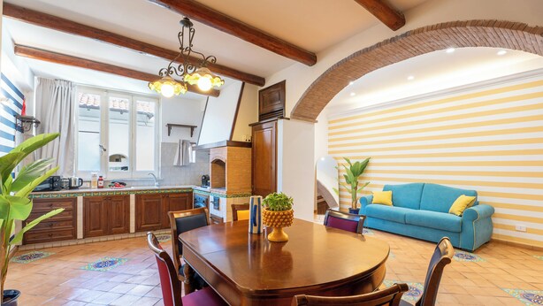 Uma zona de refeições com mesa e cadeiras de madeira, cozinha ao fundo e um sofá azul com parede listrada.