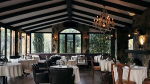 Restaurante com mesas redondas, cadeiras, um candelabro, paredes de pedra e janelas amplas.