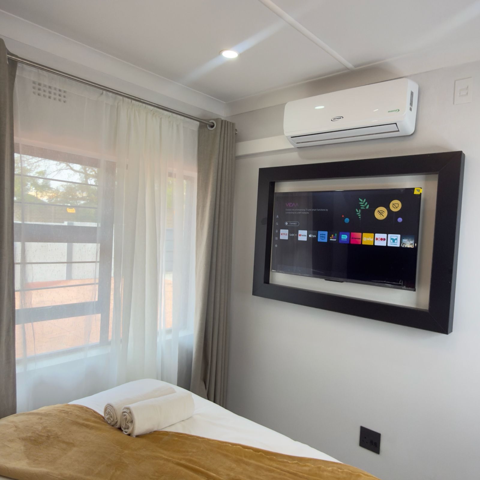 Quarto de hotel com cama, janela com cortinas, ar condicionado e televisão fixada exibindo vários aplicativos.
