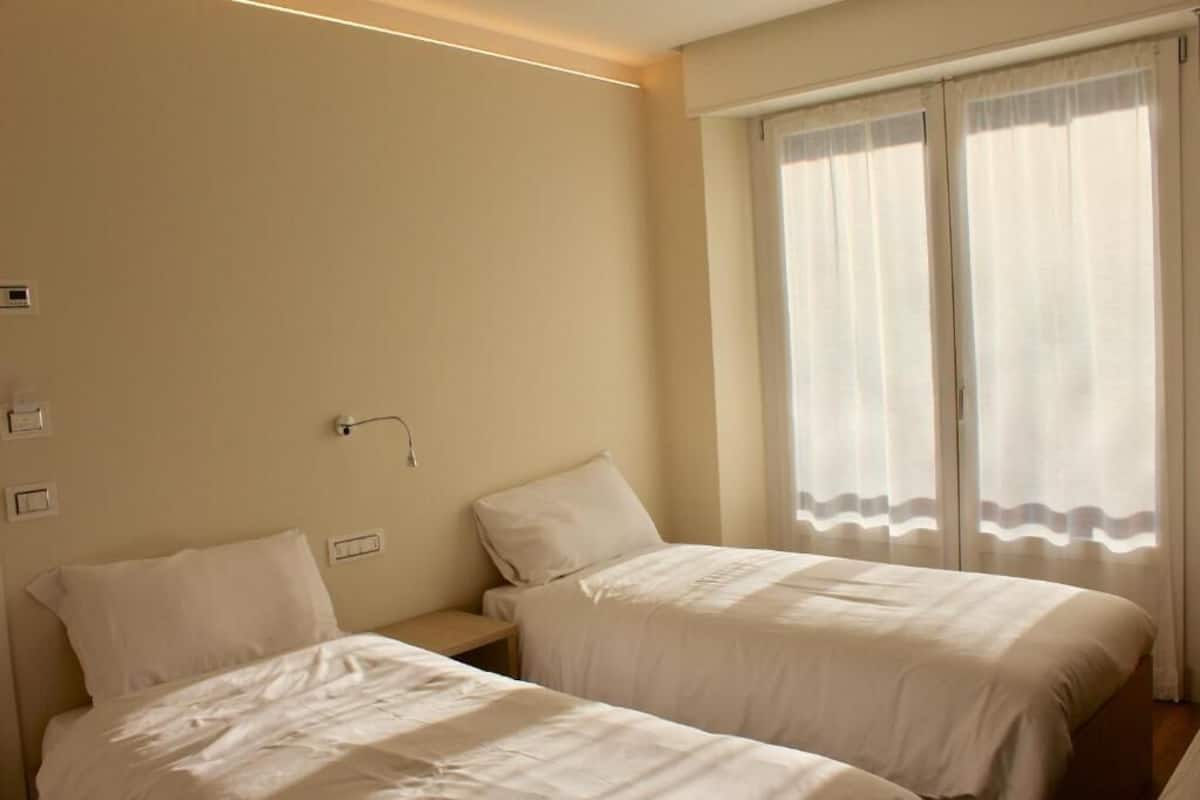 Quarto de hotel com duas camas, uma mesinha de cabeceira, uma luminária na parede e uma janela com cortinas finas.