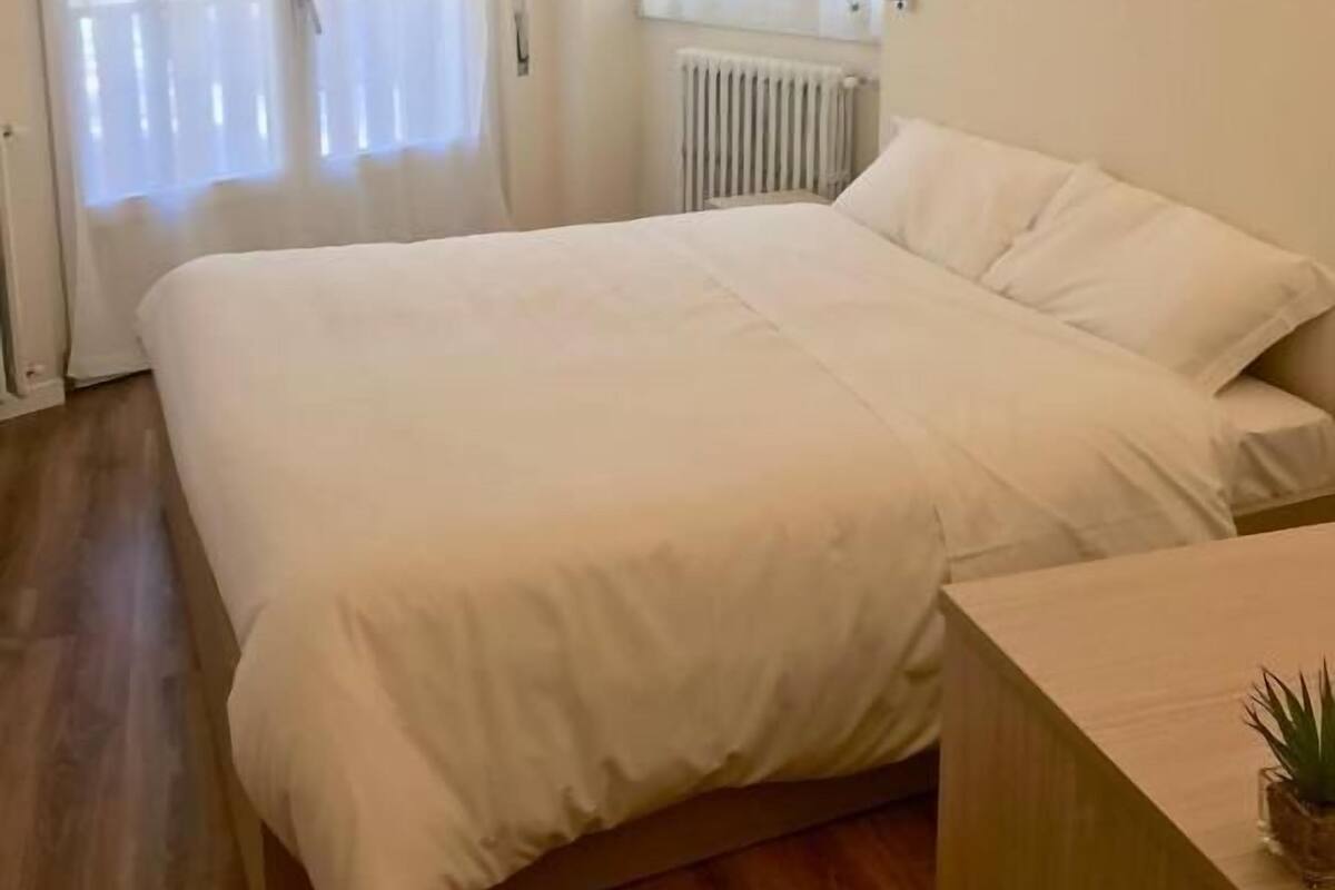 Quarto com cama, radiador e janela com cortinas.