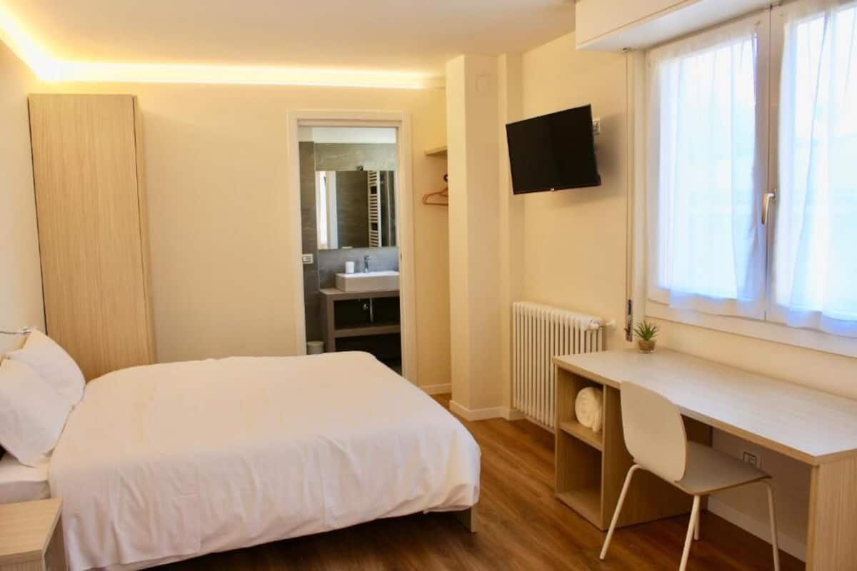 Quarto com cama, escrivaninha, televisão e janela.