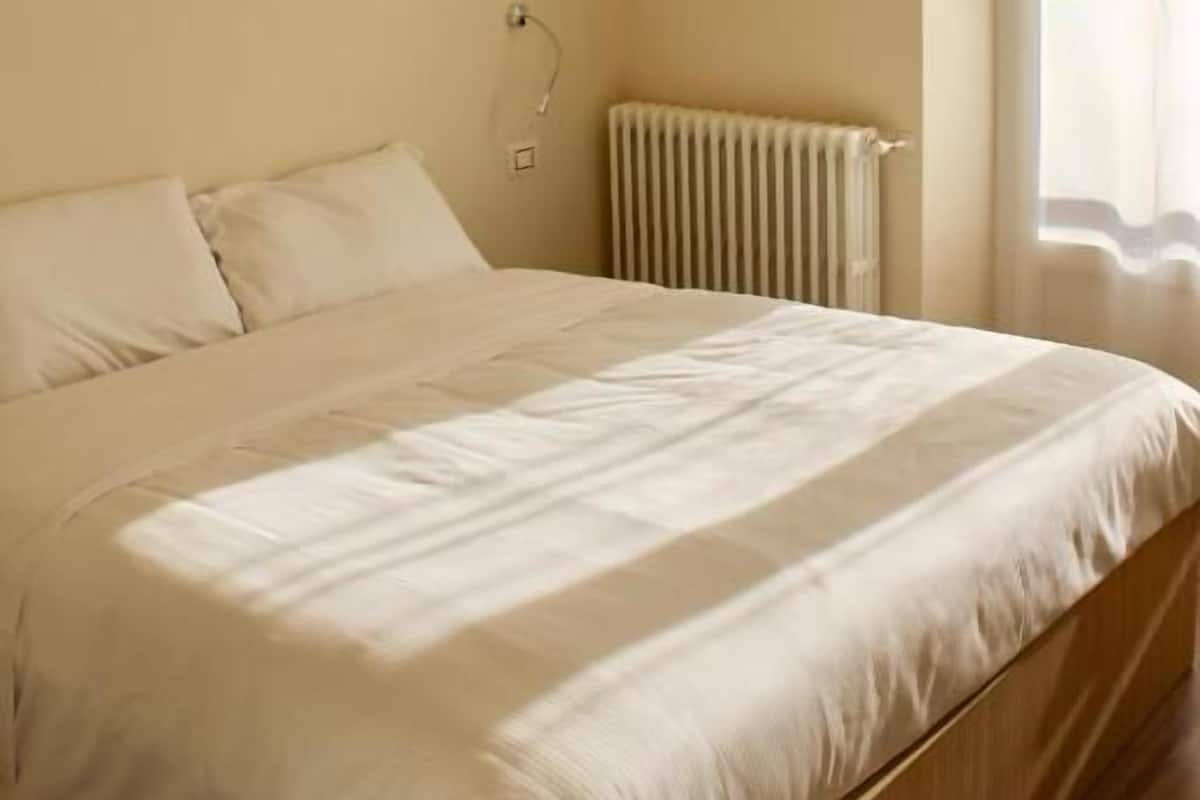 Quarto com cama, radiador, janela com cortinas e luminária fixada na parede.