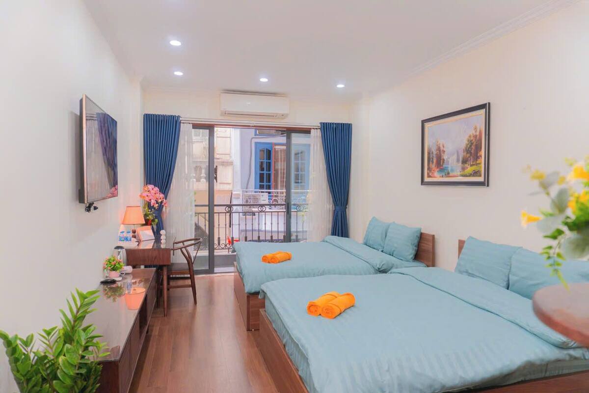 Quarto de hotel com duas camas, uma escrivaninha com cadeira, uma mesinha com abajur, uma planta em vaso e uma varanda com vista.