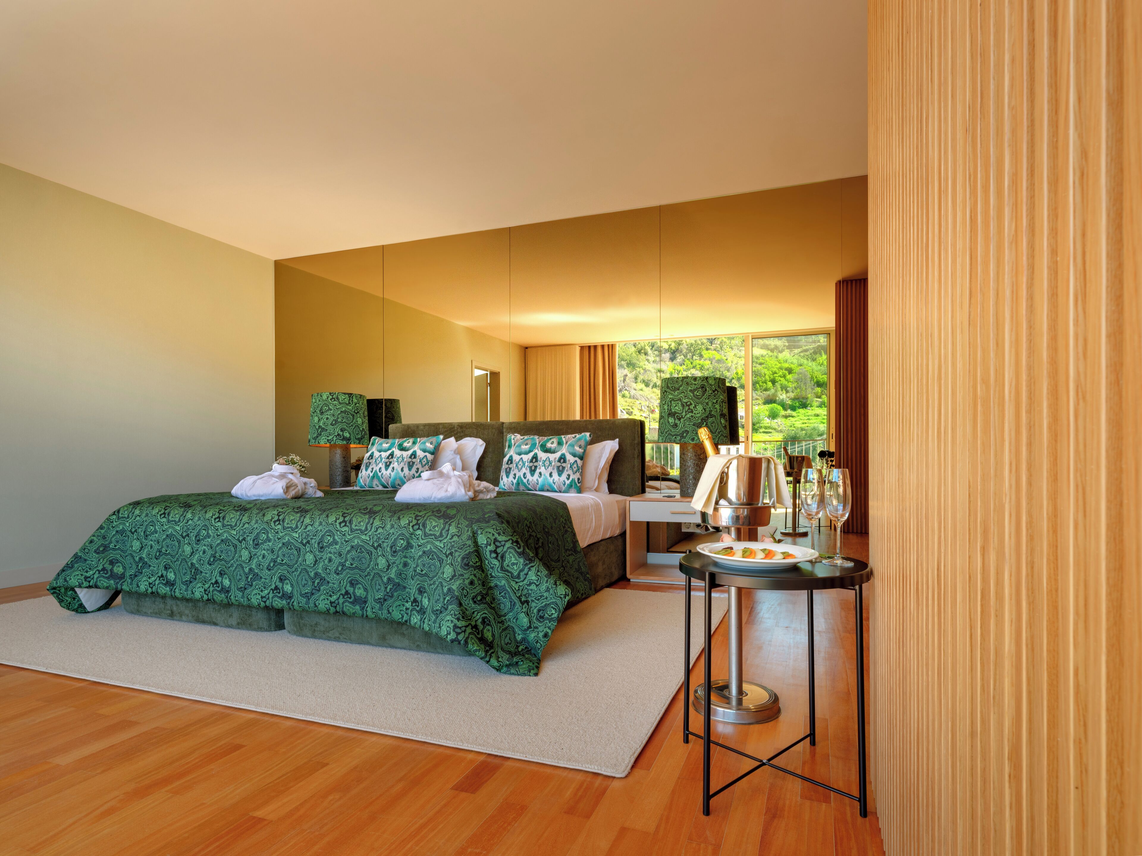 Quarto de hotel com uma cama grande, uma mesinha de cabeceira com garrafa e copos, vista para o verde lá fora e um espelho na parede.