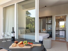 Quarto de hotel moderno com cama, banheiro e área de refeições, mesa posta com comida e vinho.