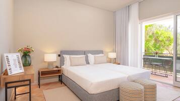 Quarto de hotel com uma cama grande, duas mesas de cabeceira com luminárias, uma pequena escrivaninha com uma foto emoldurada, um vaso com flores e uma varanda com mesa e cadeiras.
