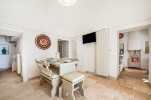 Uma sala de jantar com mesa e cadeiras, uma televisão fixada na parede e uma cozinha com uma panela pendurada.