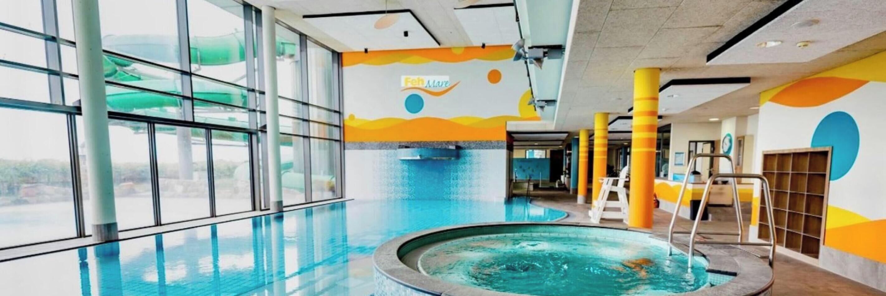 Uma piscina interior com paredes de vidro transparente, uma banheira de hidromassagem e arte colorida nas paredes.
