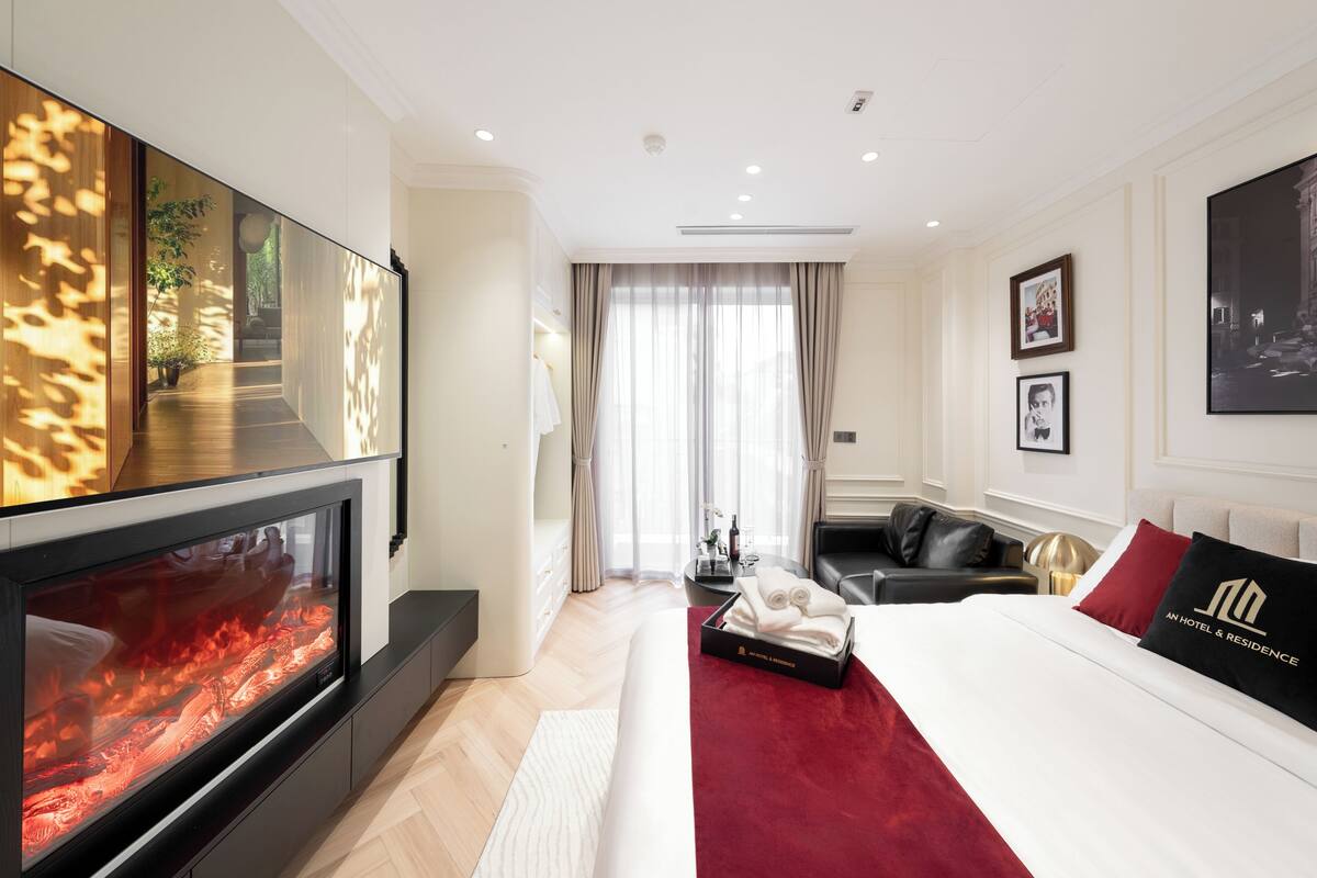 Quarto de hotel moderno com uma TV de tela plana grande, cama com manta vermelha, sofá e área de jantar com mesa e cadeiras.