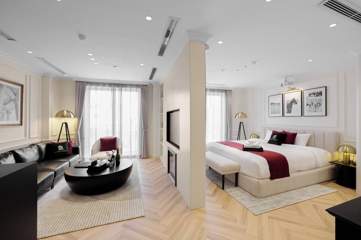 Quarto de hotel moderno com sofá, mesa de centro redonda, cama com manta vermelha, TV e quadros na parede.