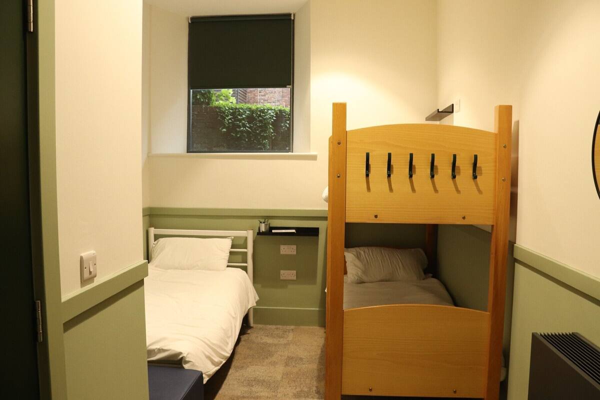 Quarto pequeno com uma cama de solteiro e uma beliche, janela com vista para o verde e radiador.