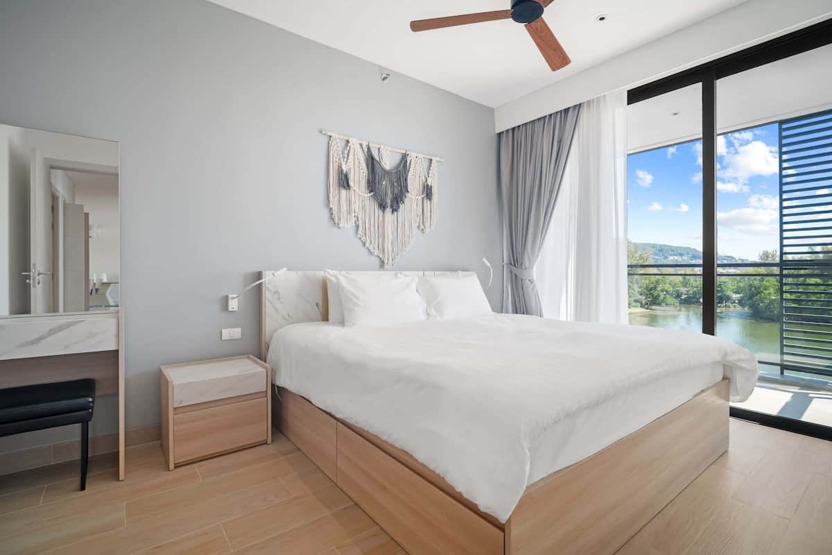 Quarto moderno com uma cama grande, mesa de cabeceira de madeira, um banco, ventoinha no teto, peça de macramé na parede e vista para um lago através de grandes janelas.