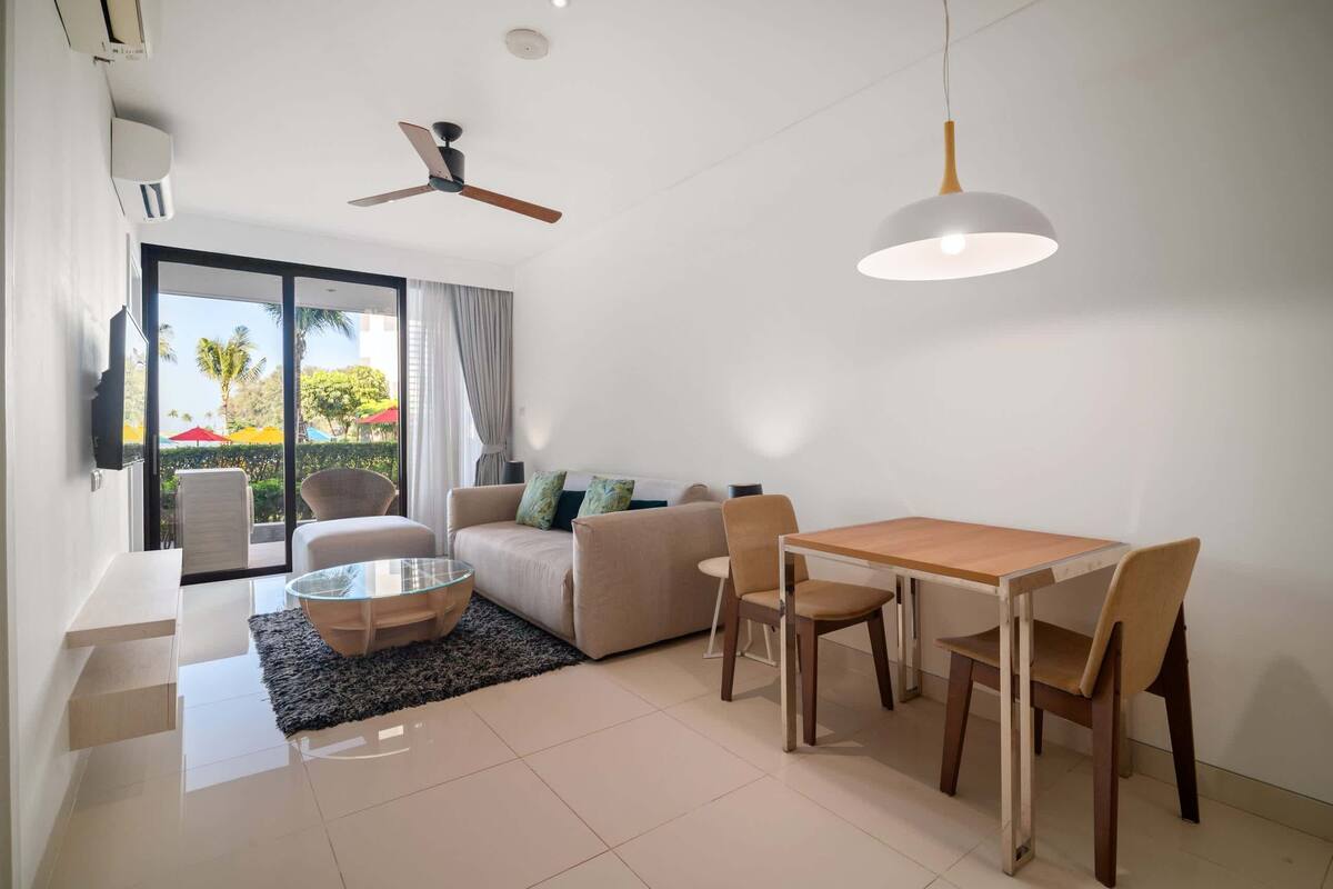 Uma sala de estar moderna com uma mesa de centro de vidro, um sofá, uma mesa de jantar com cadeiras e um ventilador de teto.