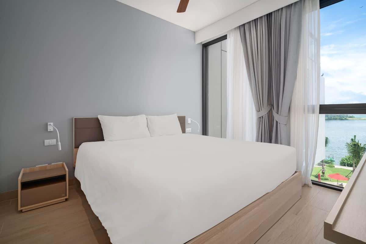 Quarto de hotel moderno com uma cama grande, uma mesa de cabeceira de madeira, uma janela com cortinas e vista para o mar.