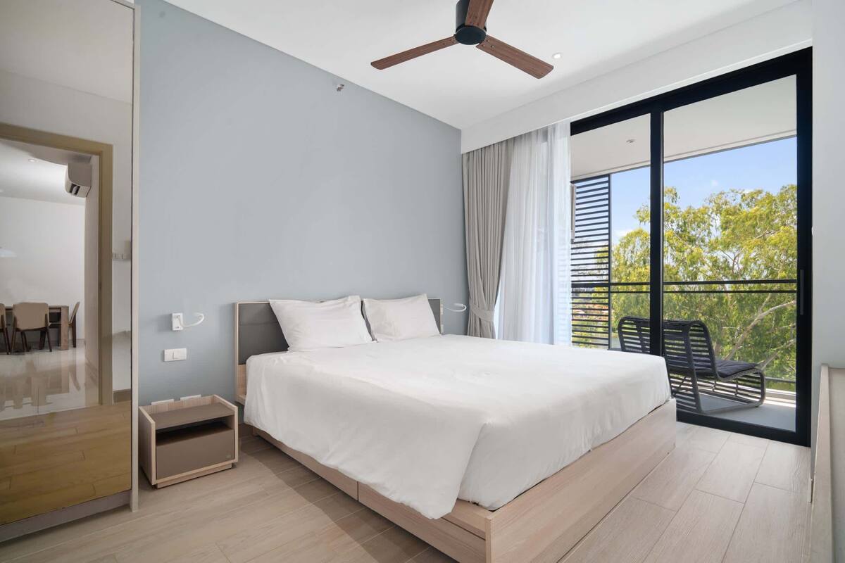 Quarto moderno com cama, ventilador de teto, varanda com cadeira e vista para árvores.