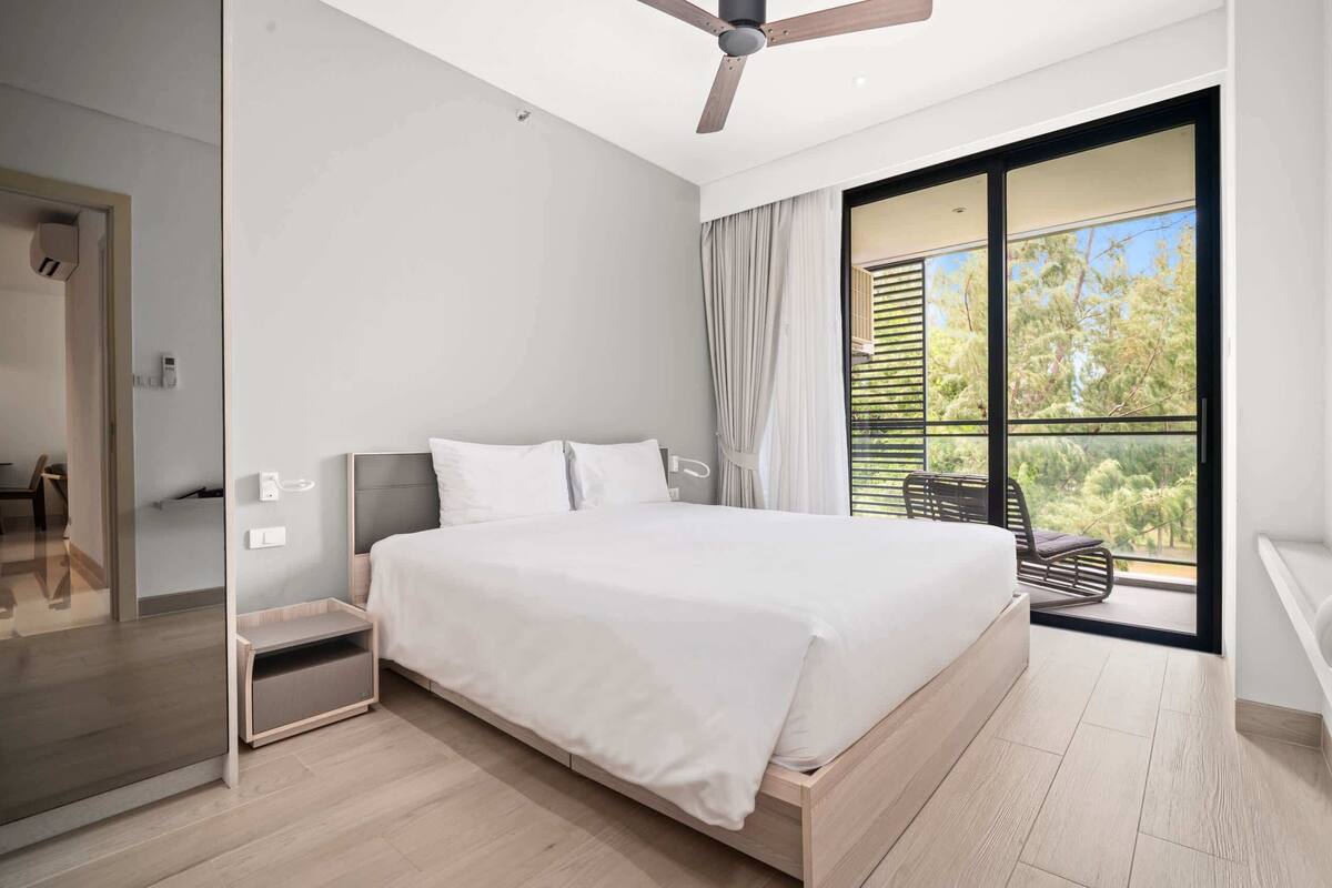 Quarto moderno com cama grande, ventilador de teto, porta de vidro deslizante para uma varanda e um banco embutido.