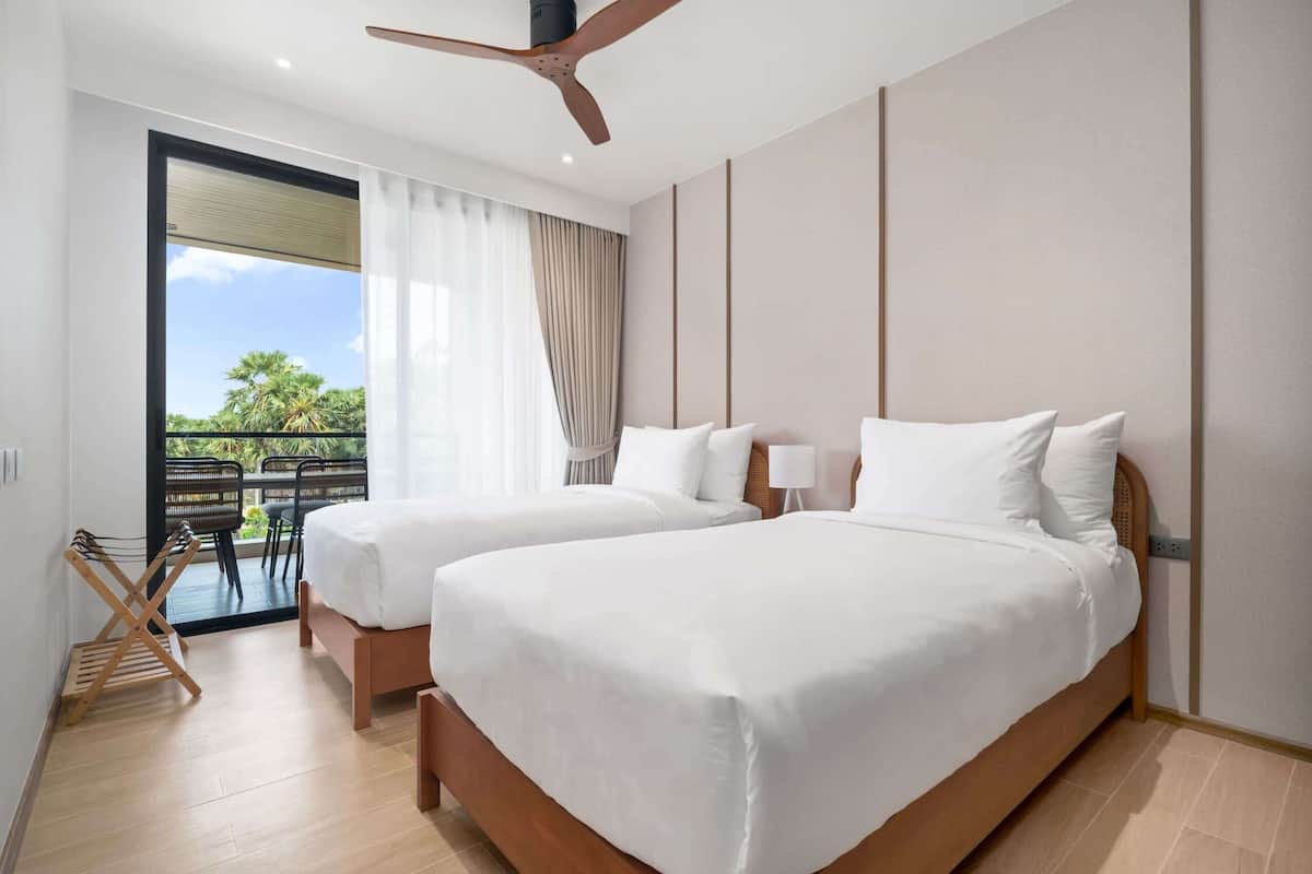 Quarto de hotel com duas camas, ventoinha no teto, varanda com vista para palmeiras e piso de madeira.