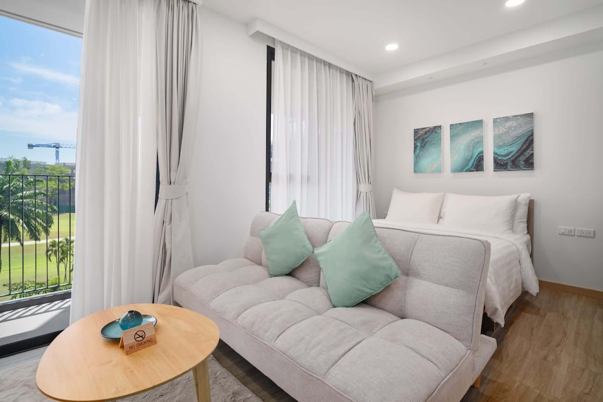 Quarto de hotel moderno com sofá, uma mesinha, cama e varanda com vista para palmeiras e uma grua de construção.