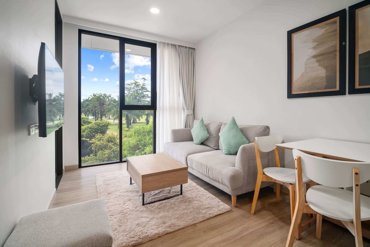 Uma sala de estar moderna com sofá, mesa de centro, mesa de jantar com cadeiras, TV fixada na parede e uma grande porta de vidro deslizante que dá para uma varanda com vista para o verde.