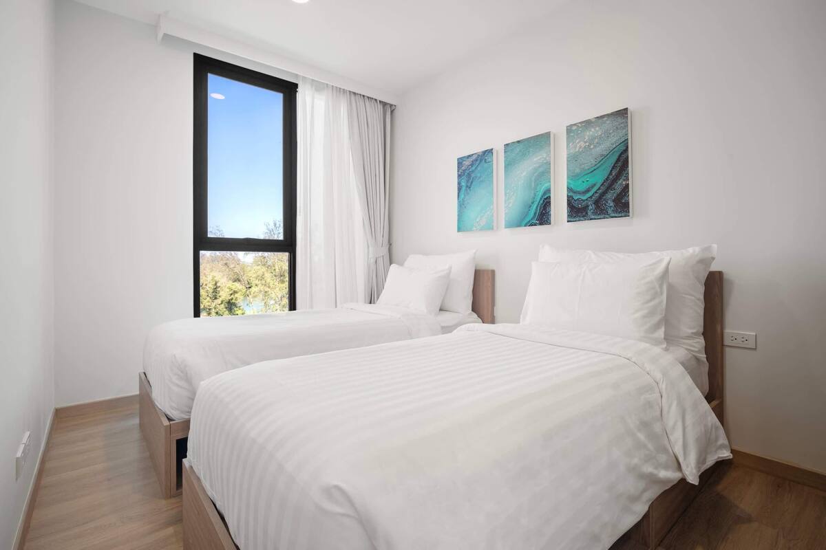 Quarto moderno com duas camas, uma janela grande com cortinas e arte abstrata na parede.