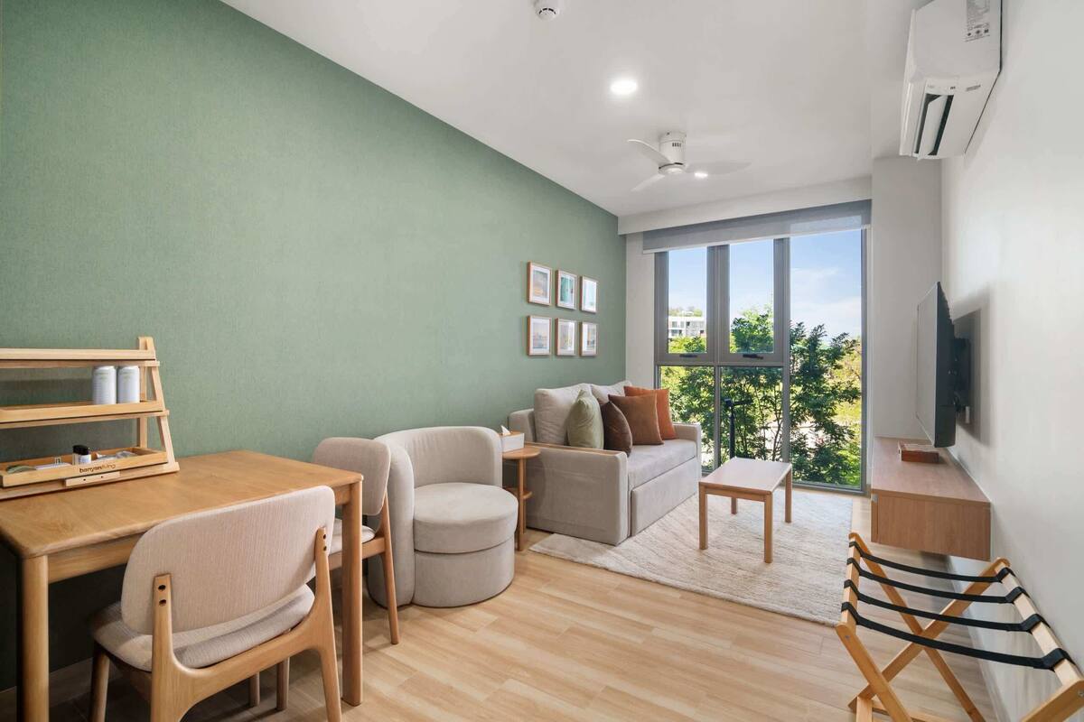 Uma sala de estar moderna com uma parede verde de destaque, um sofá, uma área de jantar com mesa e cadeiras de madeira, e uma janela grande com vista.