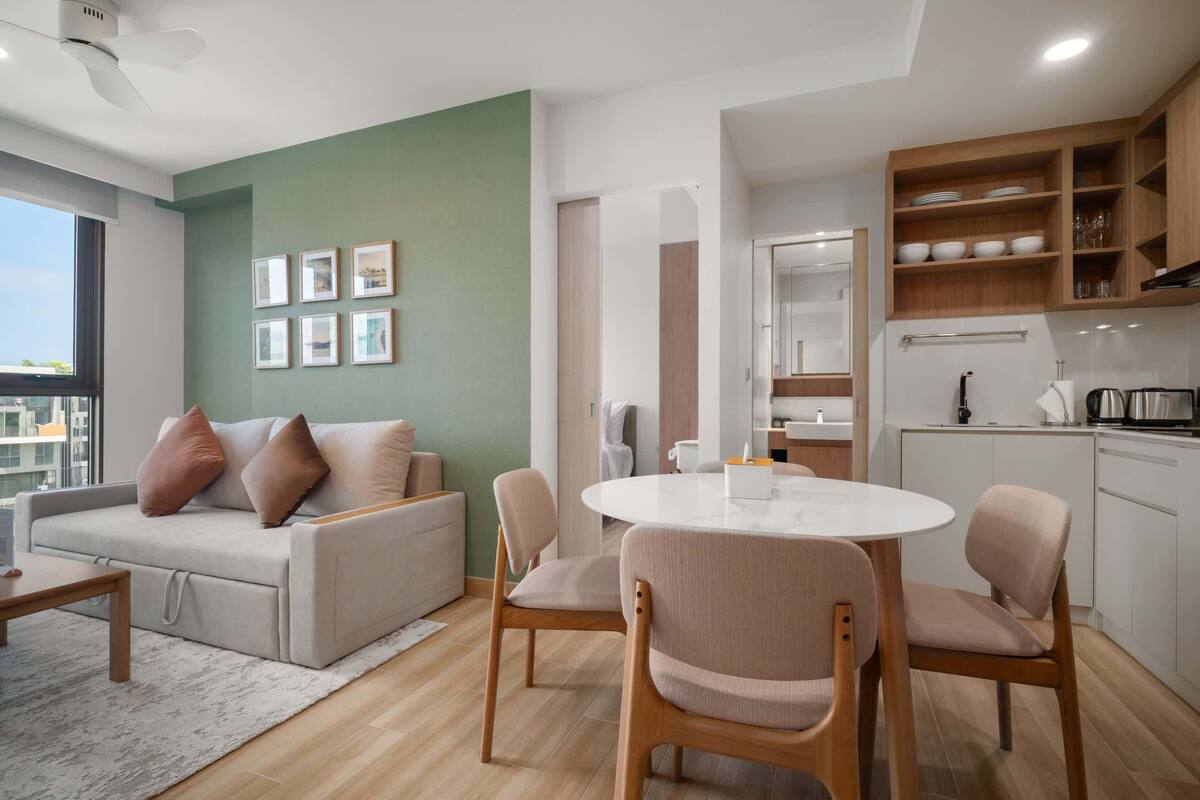 Uma sala de estar moderna com sofá, poltronas, mesa de centro e mesa de jantar. Há uma área de cozinha com armários e uma janela.