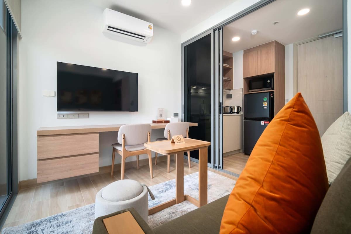 Uma sala de estar moderna com uma TV de tela plana, uma escrivaninha com cadeiras, uma mesa pequena e um sofá com um travesseiro laranja.