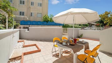 Um pátio externo com mesa, cadeiras e um guarda-sol branco.