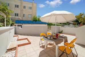 Um pátio externo com mesa, cadeiras e um guarda-sol branco.