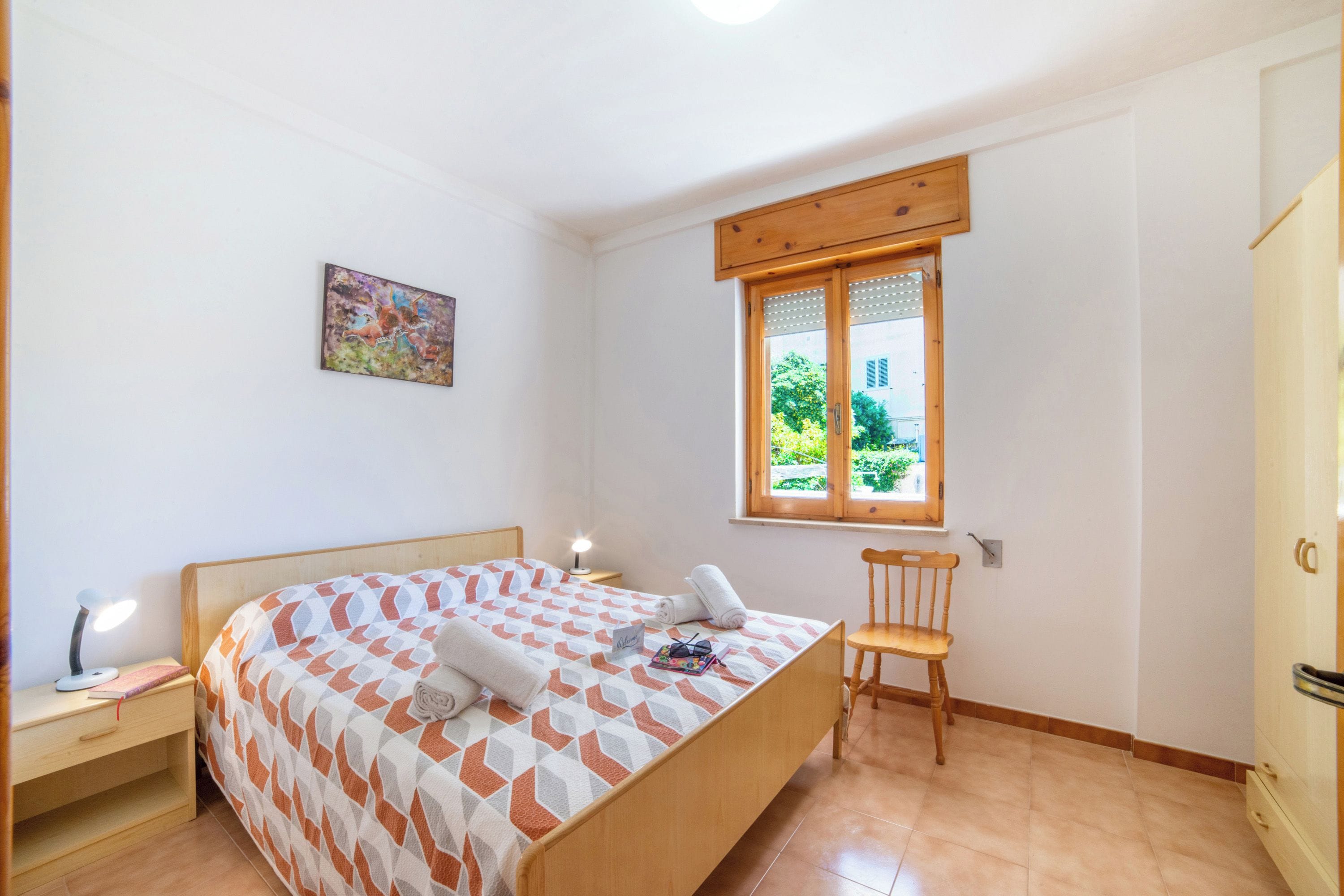 Um quarto com cama, cadeira, escrivaninha, janela e um quadro na parede.