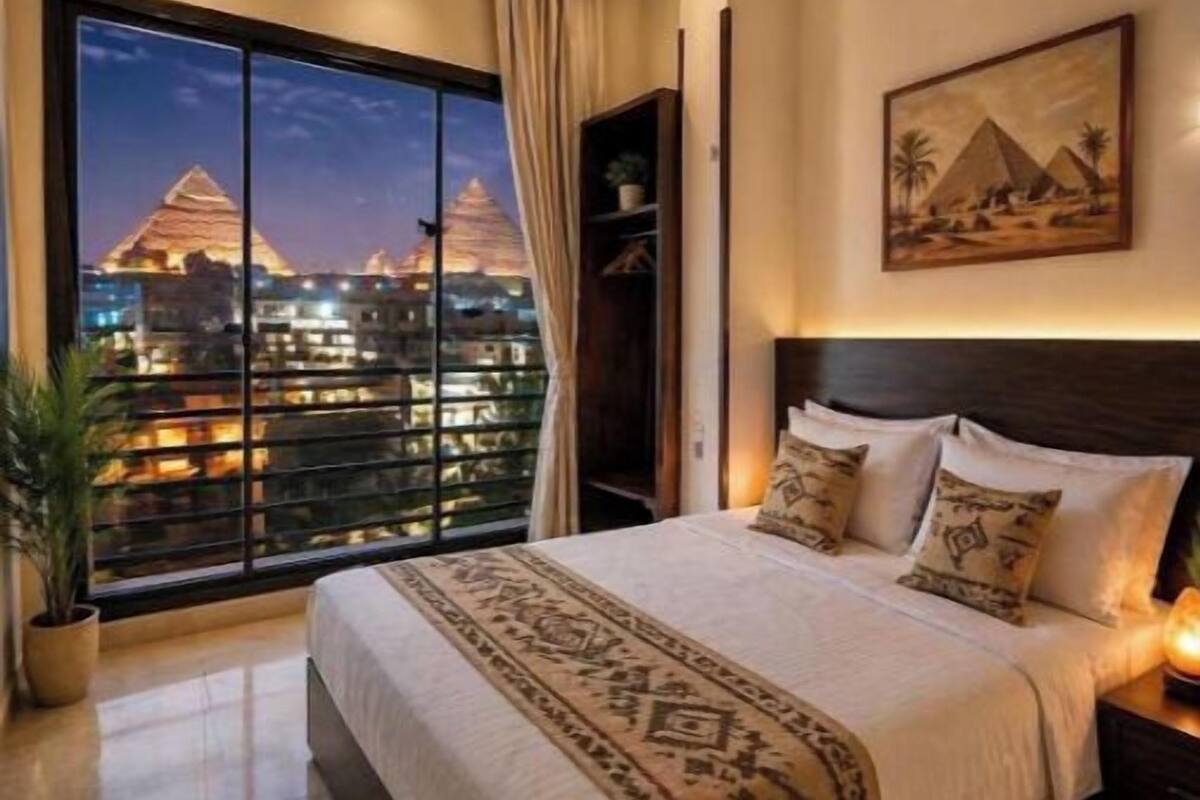 Quarto com uma cama grande, mesa de cabeceira, luminária, pintura, janela com vista para pirâmides e uma planta em vaso.